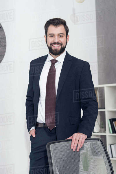 Portrait of cheerful businessman in suit standing at workplace in ...