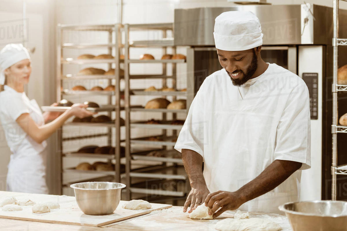 Handsome african american baker kneading dough while his female ...