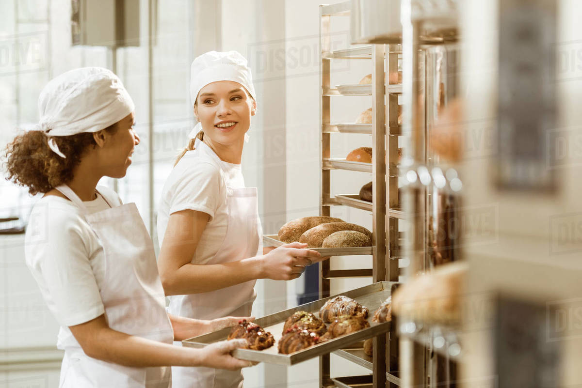 Attractive female bakers putting fresh pastry on shelves at baking ...