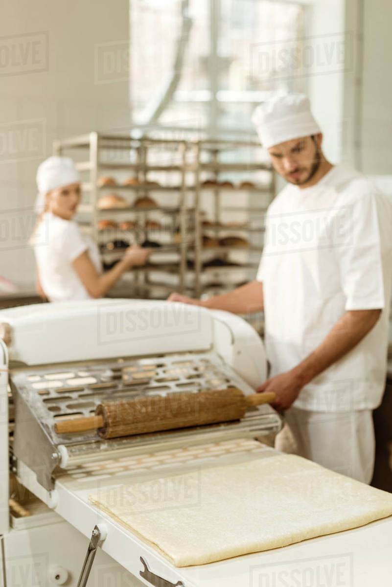 Handsome young baker working with industrial dough roller at baking ...