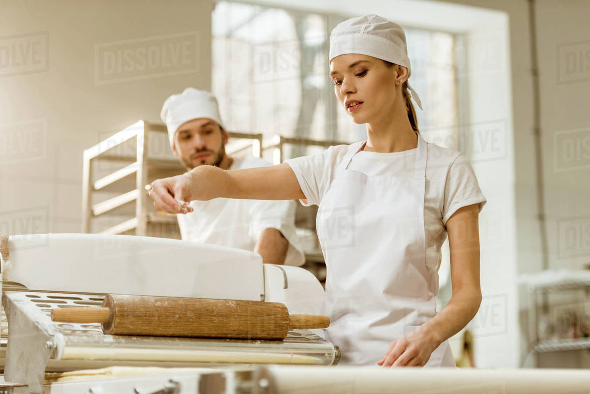 Young bakers in uniform working with industrial dough roller at baking ...