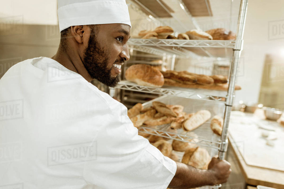 Happy african american baker driving shelves of fresh bread on baking ...