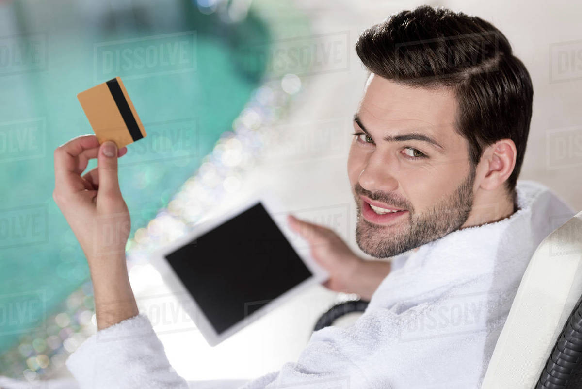 Smiling young man in bathrobe holding credit card and digital tablet in ...
