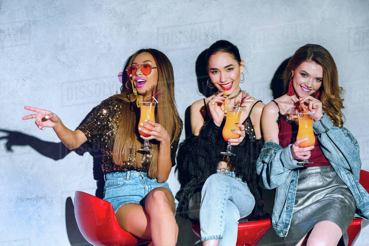 Happy stylish multiethnic girls sitting on bar stools and drinking ...