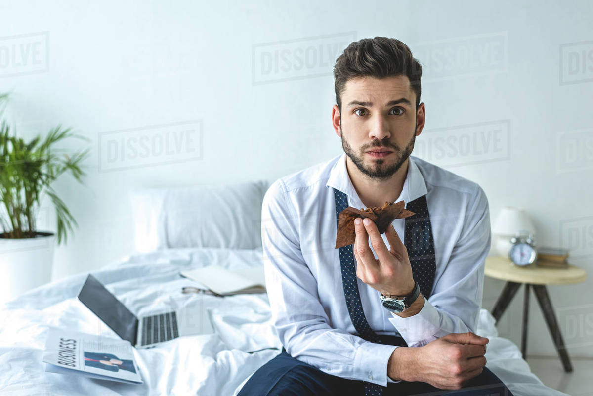 Businessman eating muffin on bed with laptop and newspaper - Royalty ...