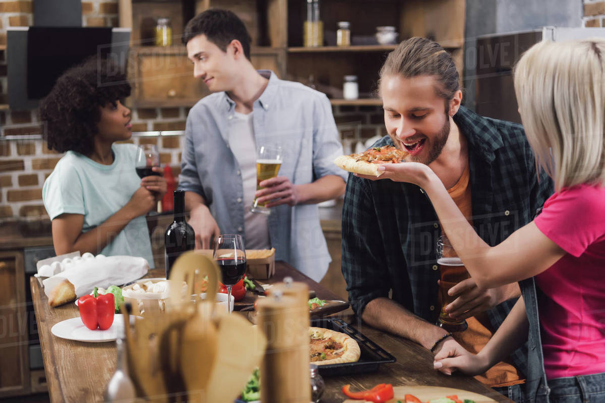 Girl feeding friend with piece of pizza in kitchen - Royalty-free Stock ...