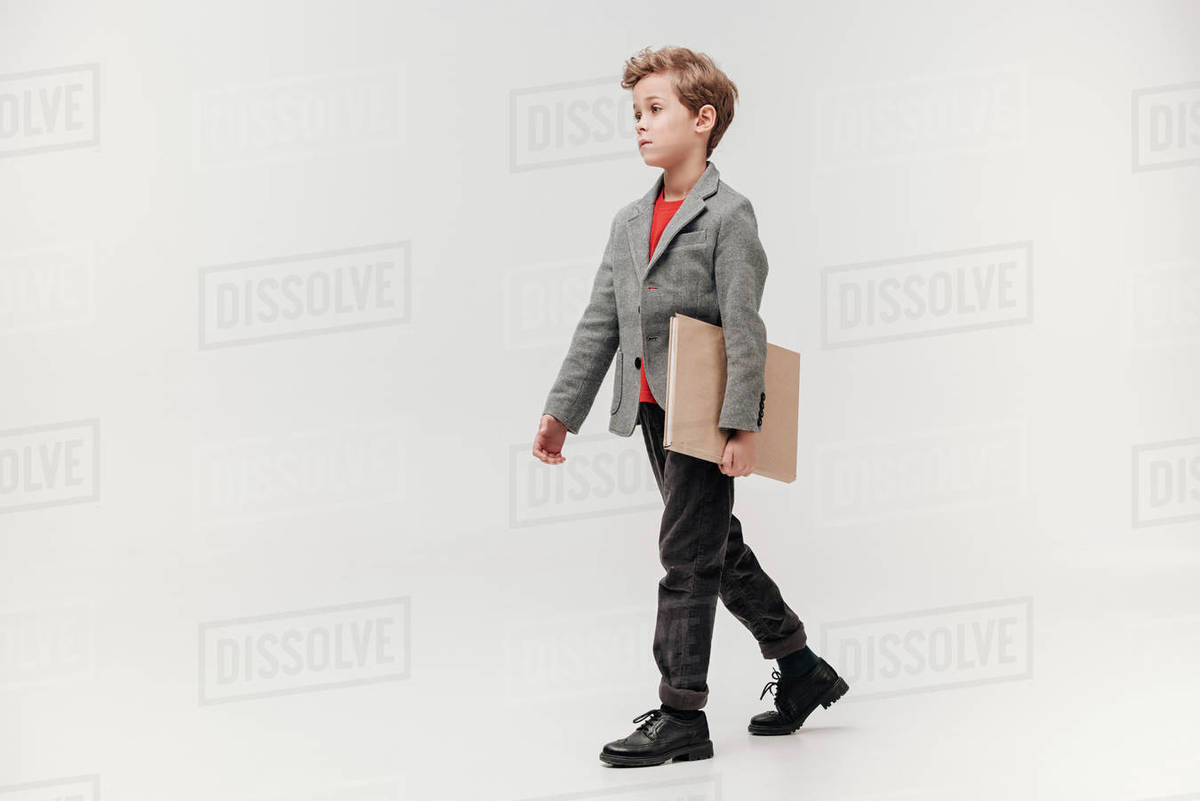 stylish little schoolboy walking with big book isolated on grey ...