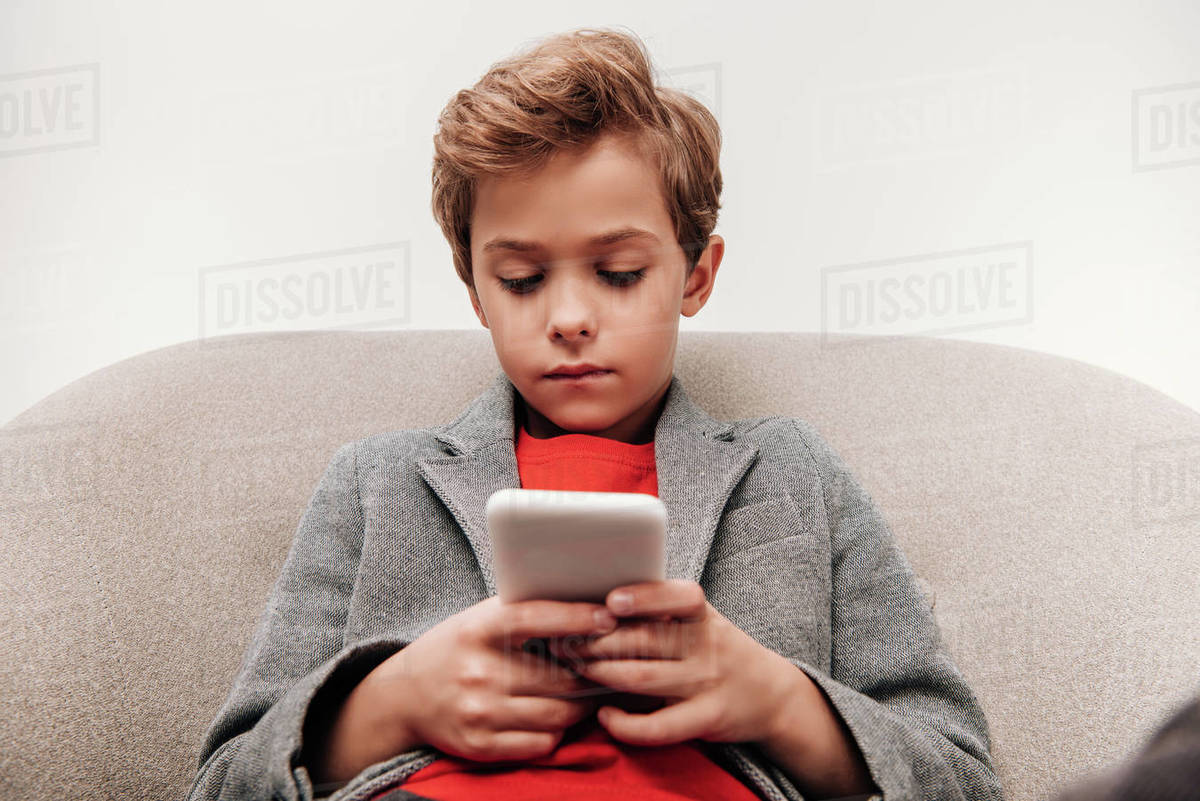 serious little boy using smartphone while sitting in armchair isolated ...