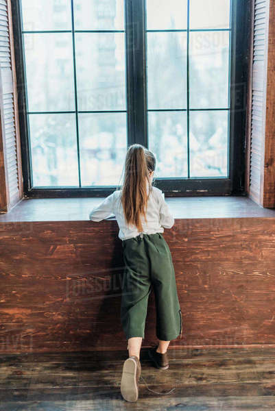 Back view of little child in stylish clothing standing at window ...