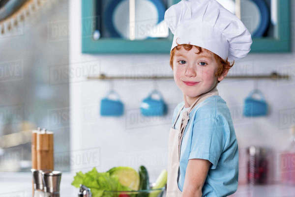 Cute little boy in chef hat and apron smiling at camera in kitchen ...