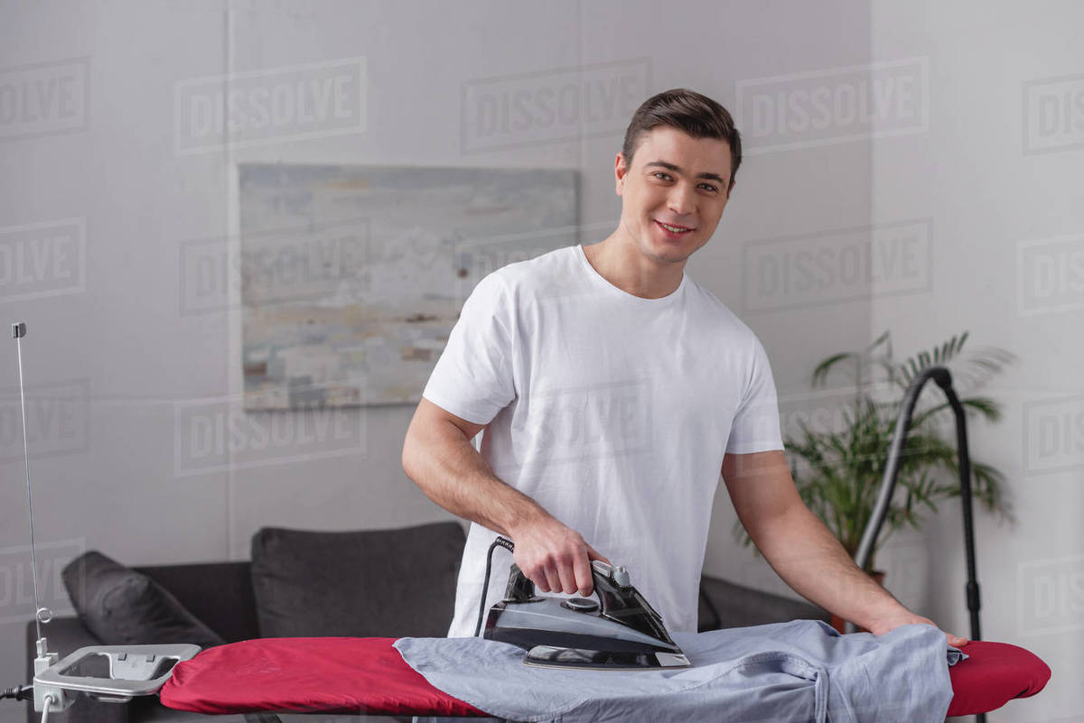 Smiling handsome man ironing shirt on ironing board in living room ...