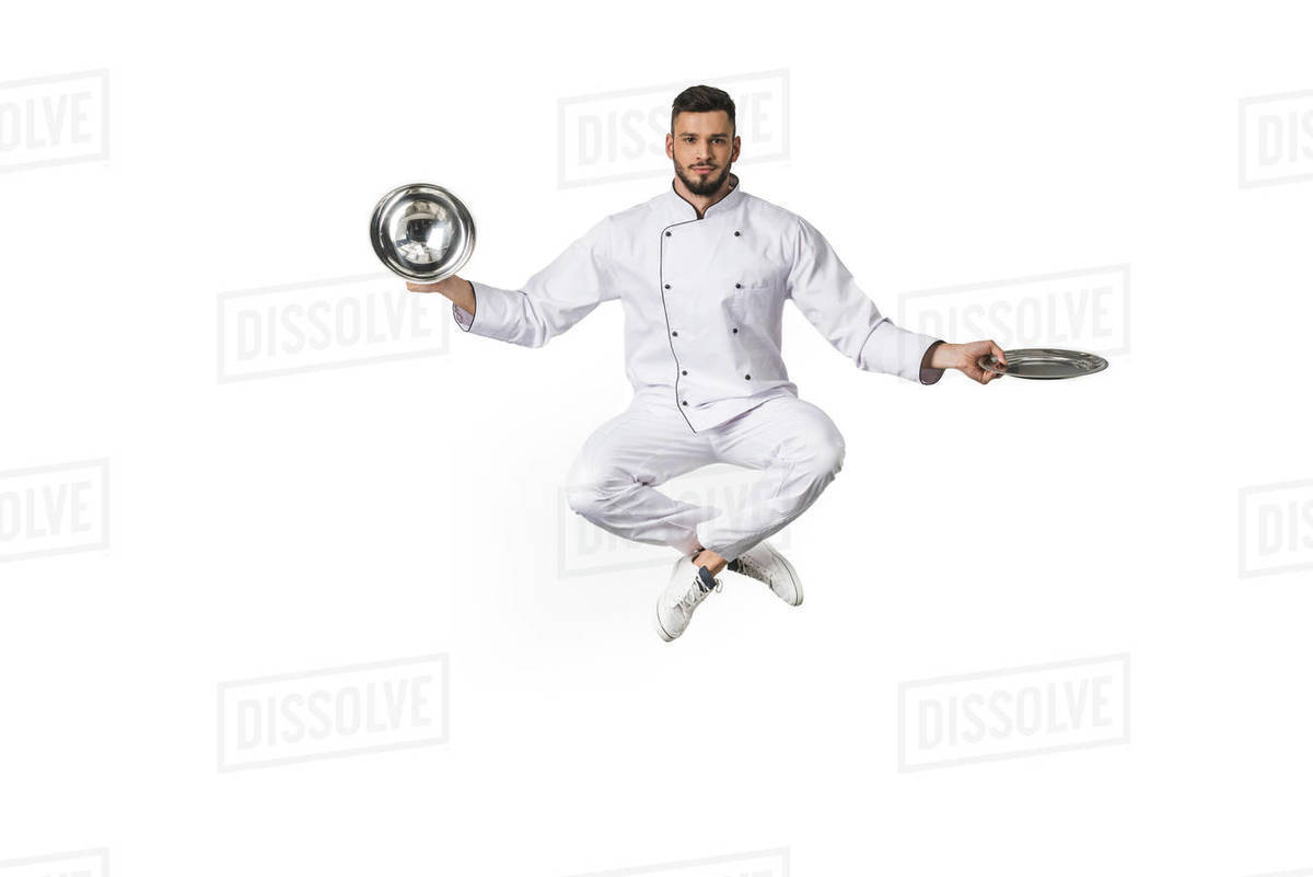 Young male chef with plates jumping and looking at camera isolated on ...
