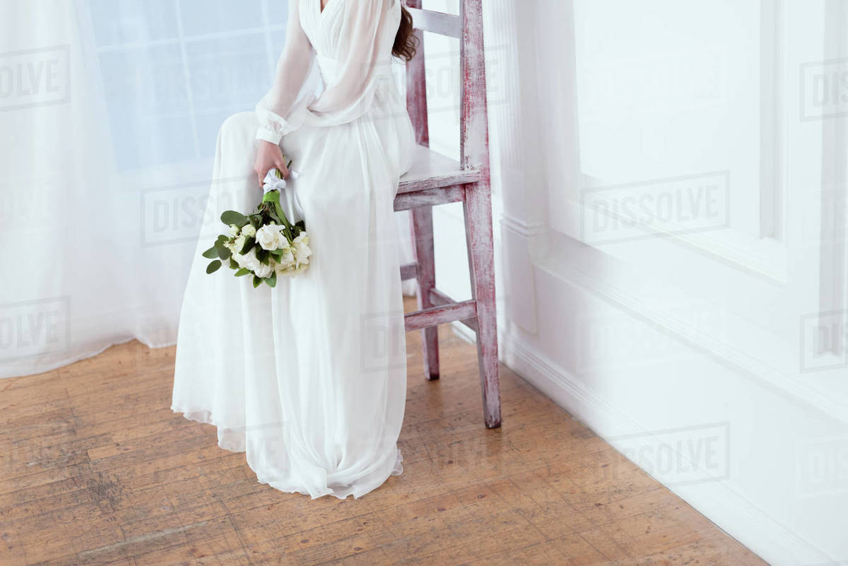 Cropped view of elegant bride sitting on big chair with wedding bouquet ...