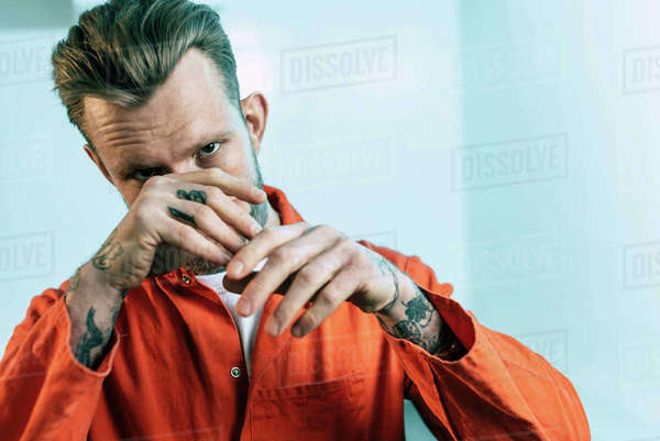 prisoner with tattoos covering face with hands and looking at camera ...