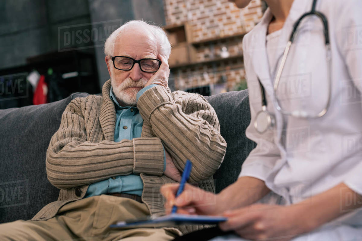 Sad senior man looking at doctor writing down medical complaints ...