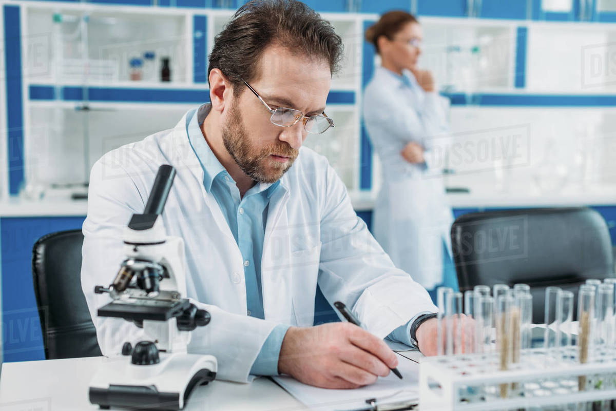 selective focus of scientist in white coat and eyeglasses making notes ...
