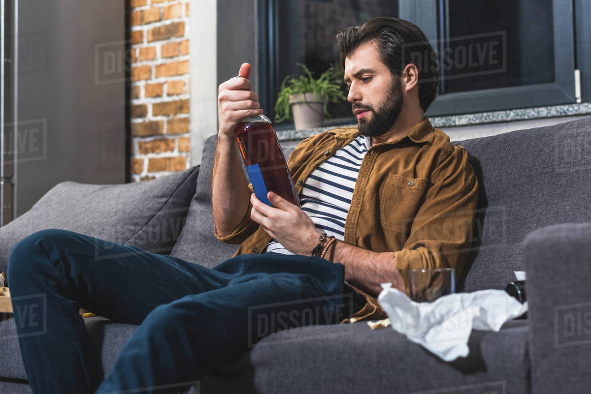 Handsome loner looking at bottle of alcohol drink at living room ...
