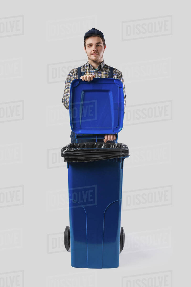 Portrait of young cleaner in uniform with trash bin isolated on grey ...