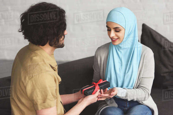 Muslim boyfriend presenting gift box to girlfriend in hijab in living ...