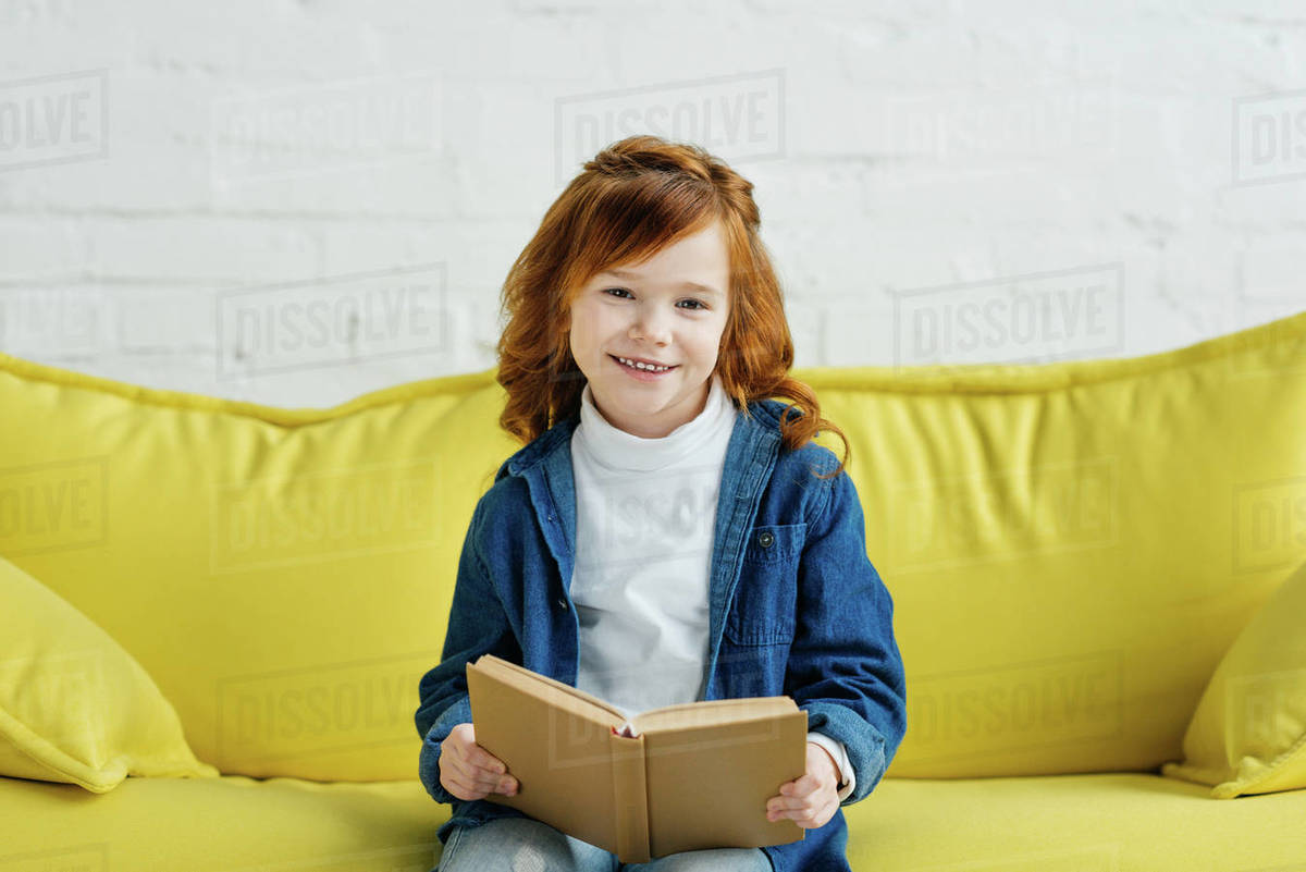 Smiling little child holding book on sofa - Stock Photo - Dissolve