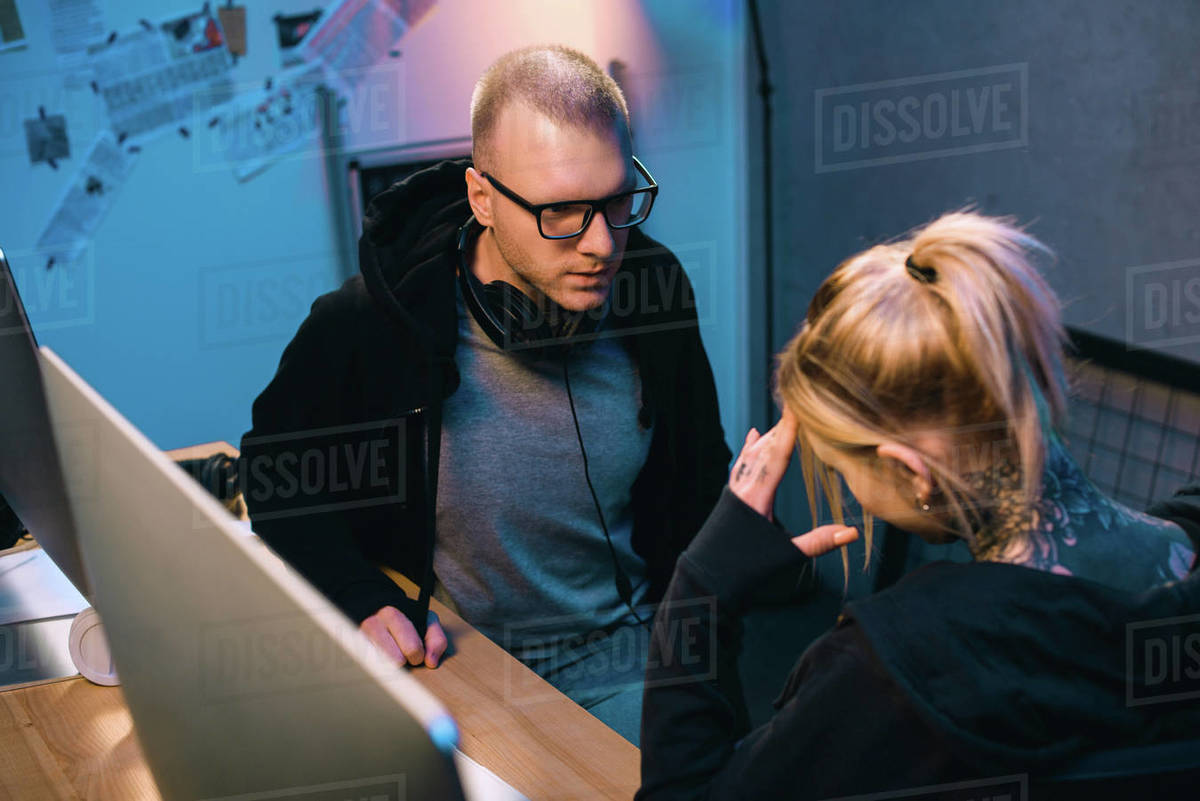 Young hacker talking with depressed accomplice at workplace in dark ...