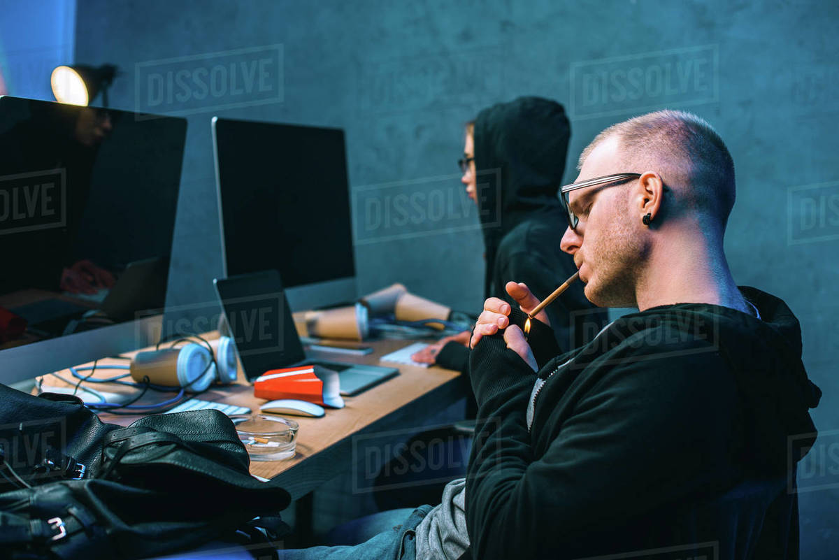 Side view of hacker working on malware with accomplice and smoking ...