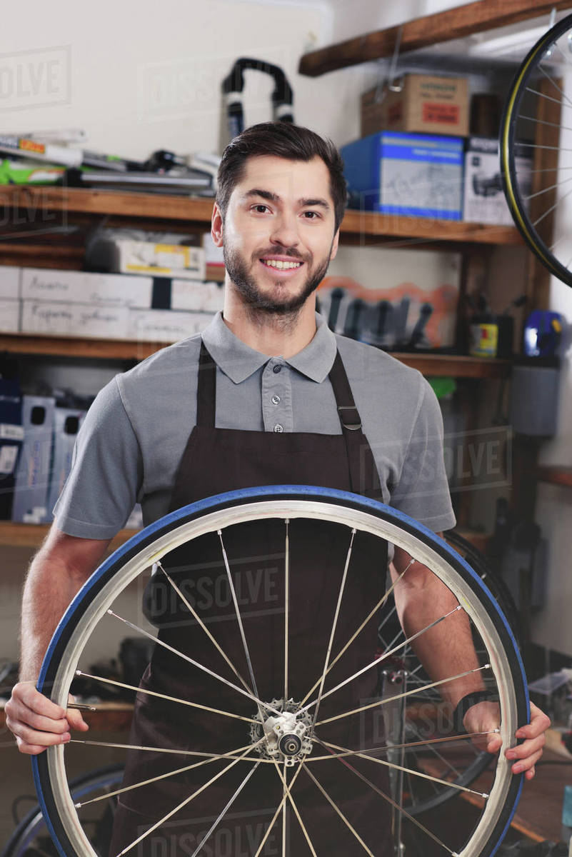 Handsome young worker in apron holding bicycle wheel and smiling at ...