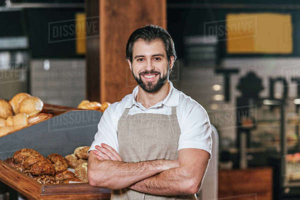 Portrait of smiling shop assistant in apron with arms crossed looking ...