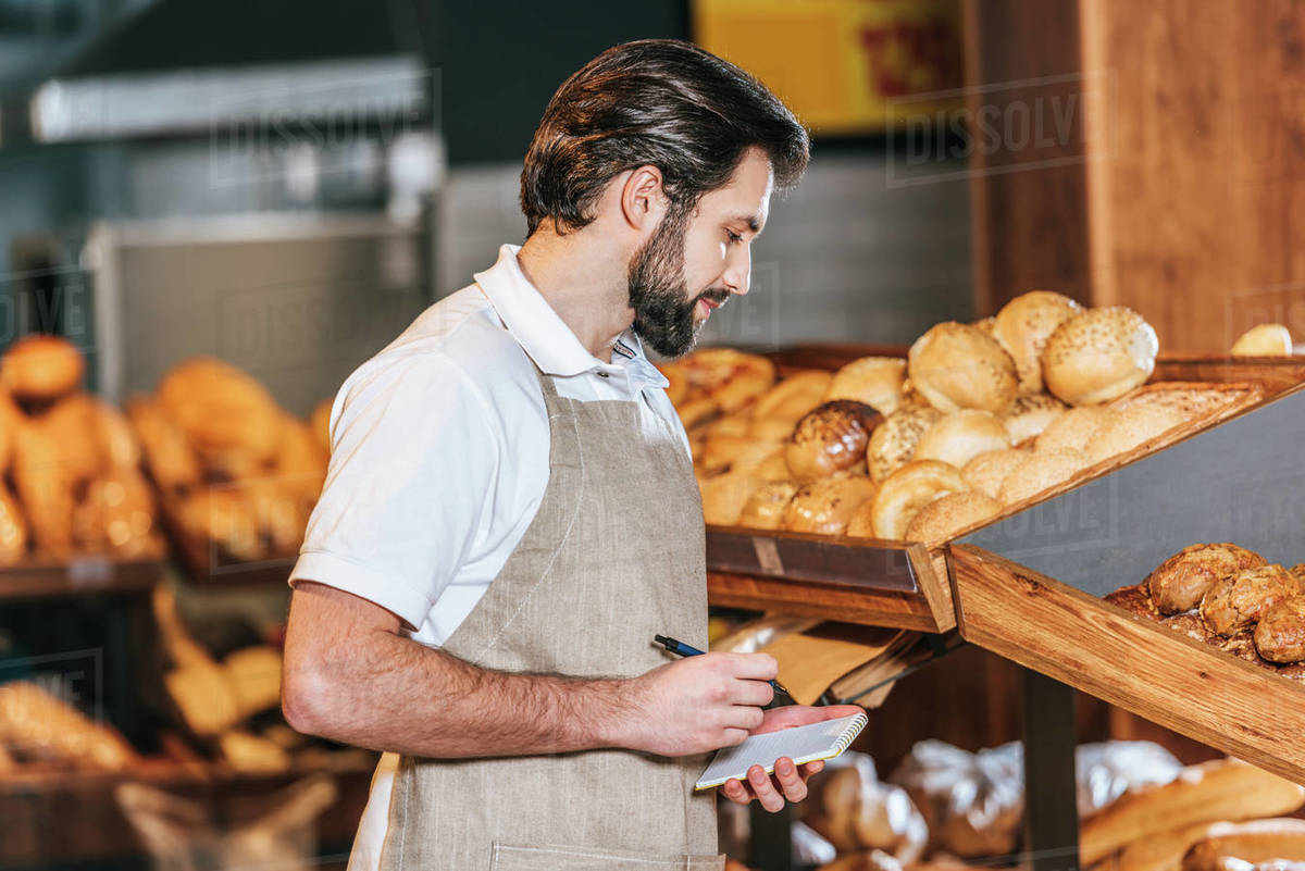 Male shop assistant in apron making notes in notebook in supermarket ...