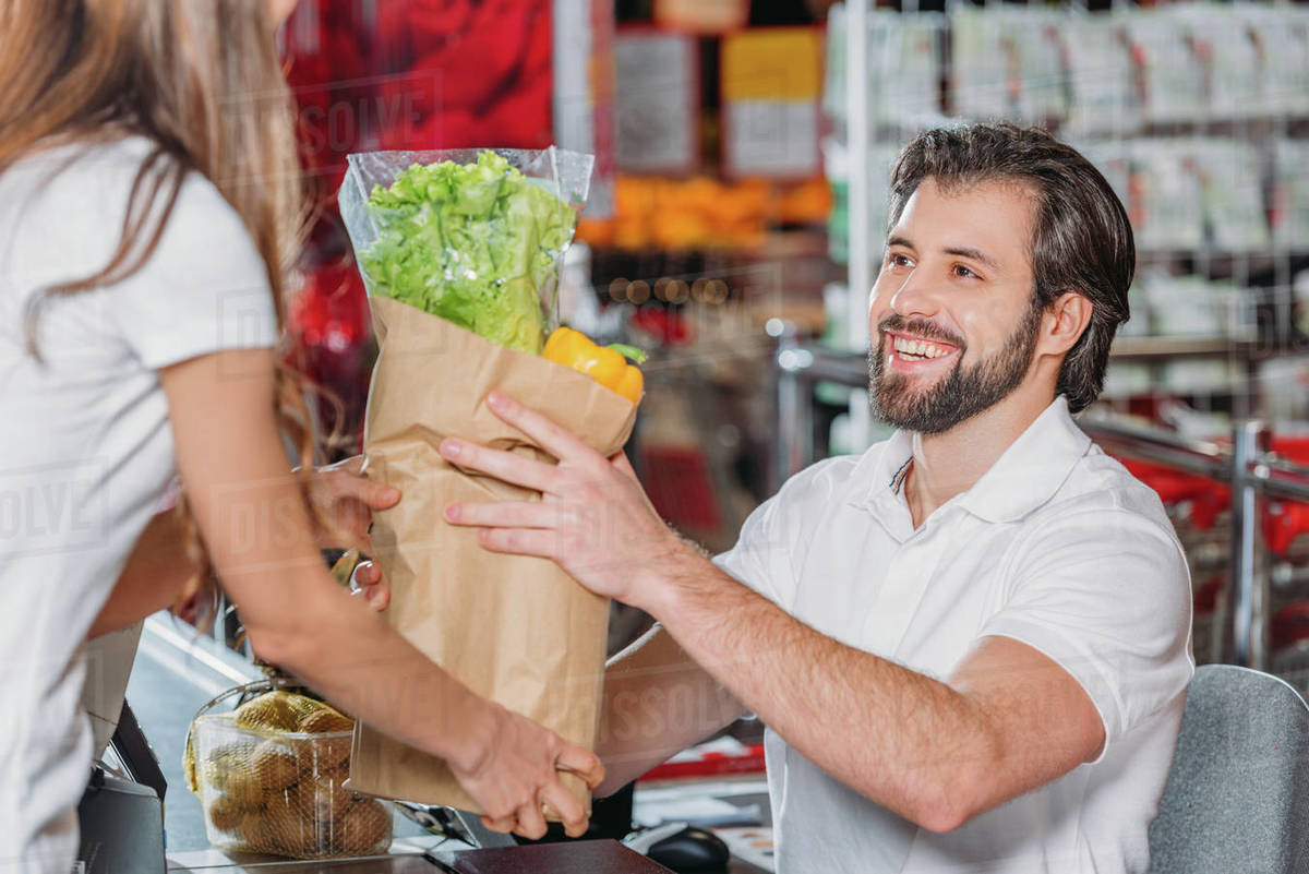 Smiling shop assistant giving purchase to shopper in supermarket ...