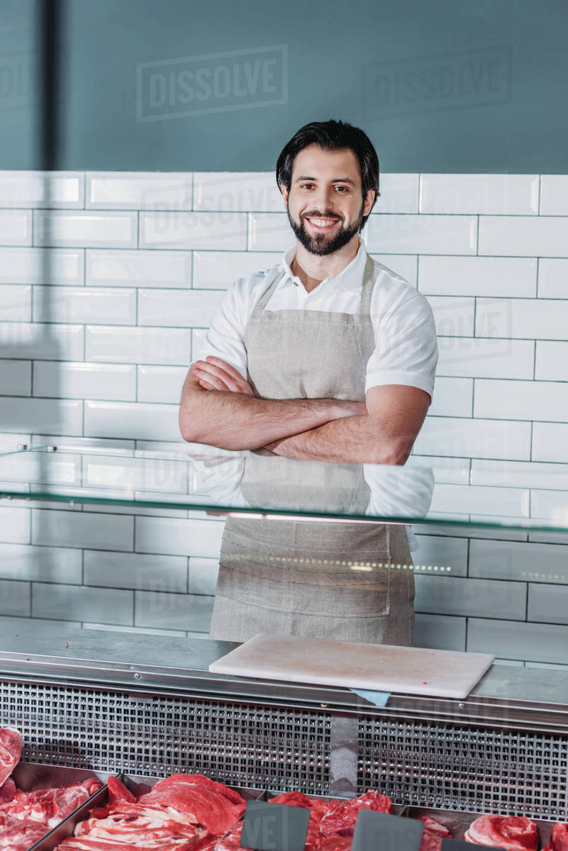 Portrait of smiling shop assistant in apron with arms crossed standing ...