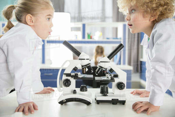 Schoolchildren in lab coats using microscopes and looking at each other ...