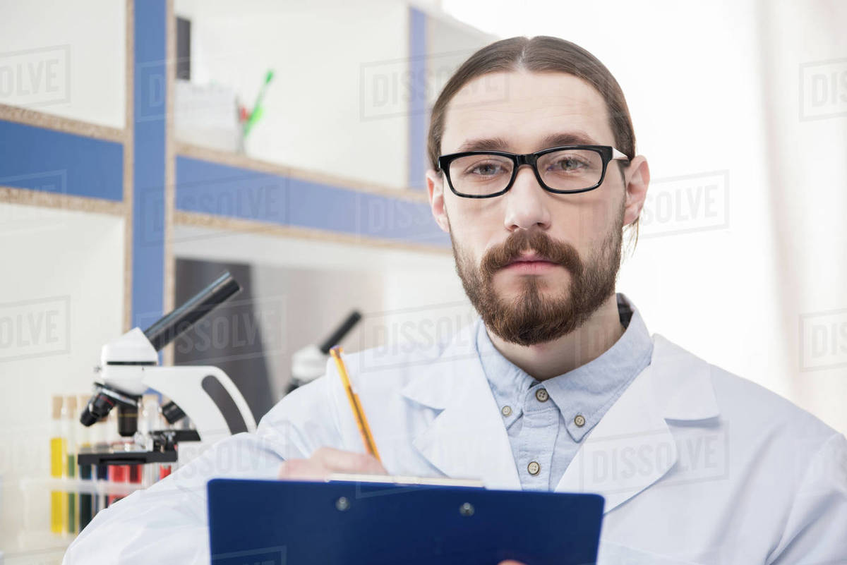Young male scientist in eyeglasses making notes and looking at camera ...