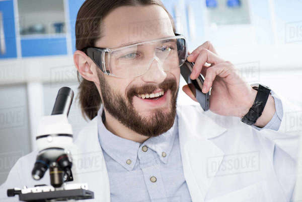 Happy young male scientist in protective glasses talking on smartphone ...