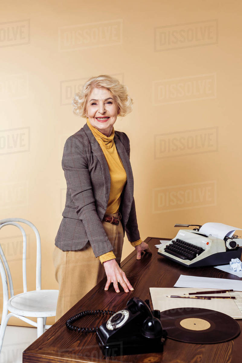Stylish senior woman standing at table with rotary phone, vinyl plate ...