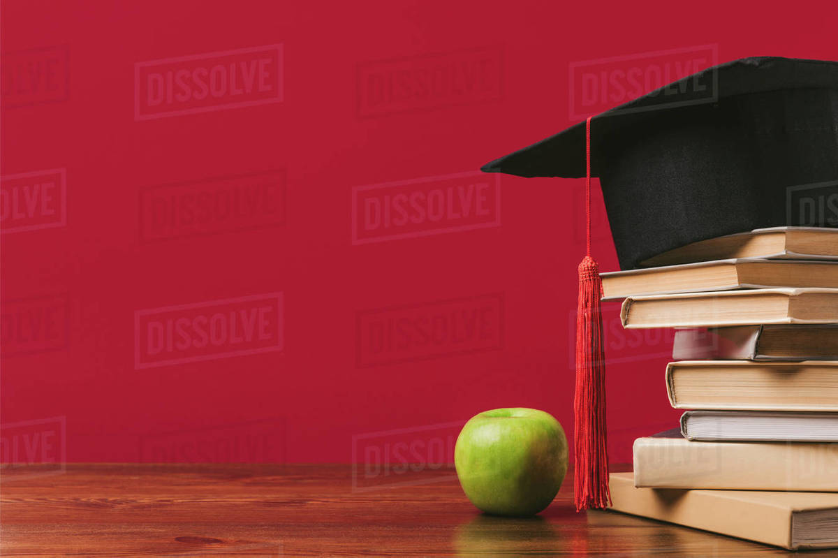 Cropped view of pile of books with academic cap on top and apple on red ...