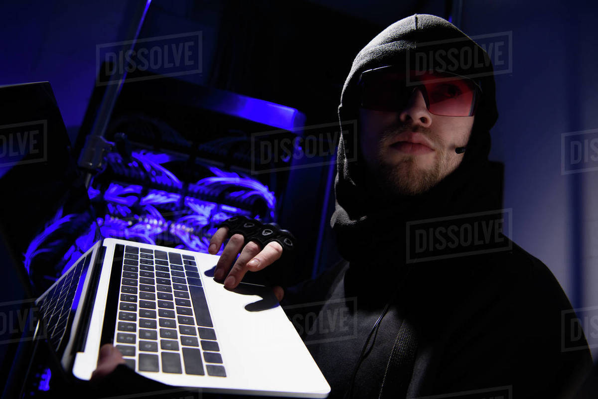 Side view of hacker in eyeglasses with laptop looking away with cables ...