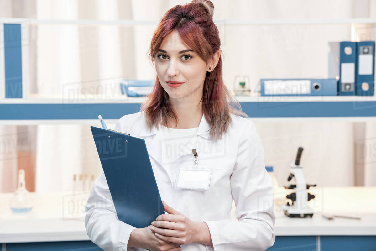 young scientist in lab coat holding folder and looking at camera in ...