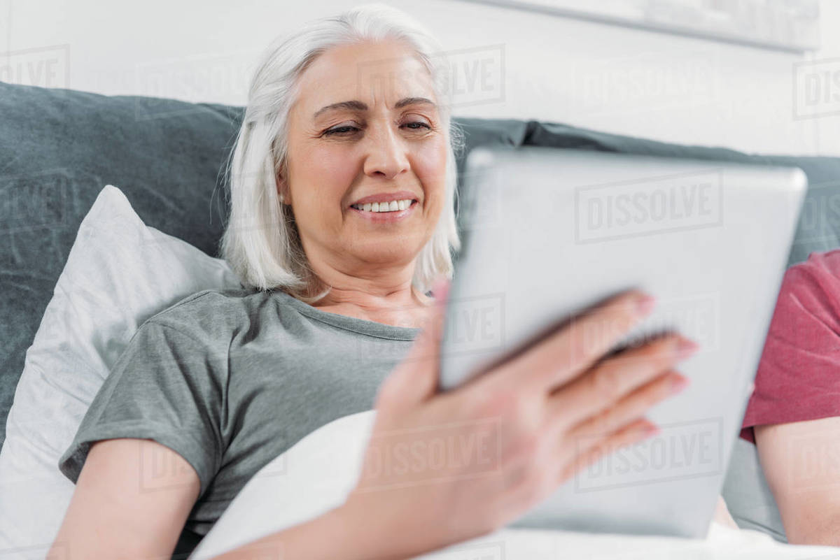 Portrait of smiling woman using tablet while lying in bed at home ...