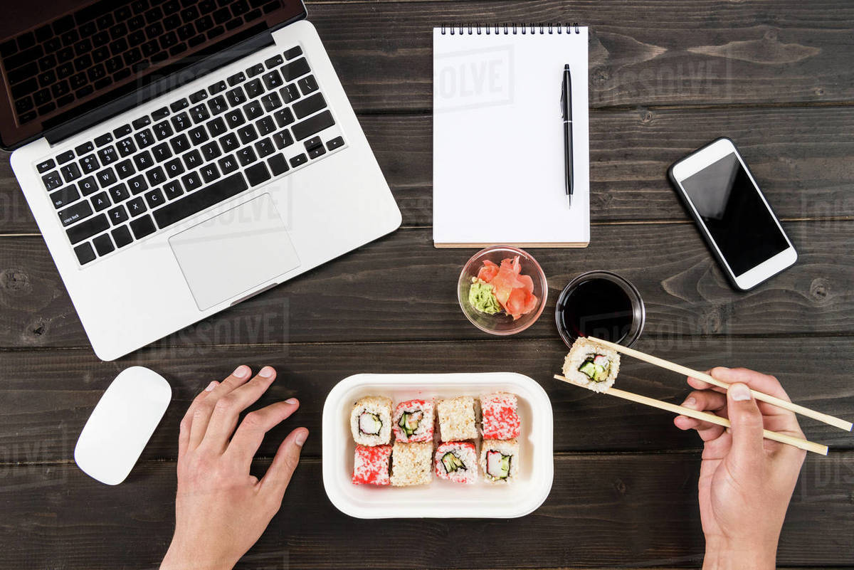 Top view of human hands eating sushi during work with digital devices ...