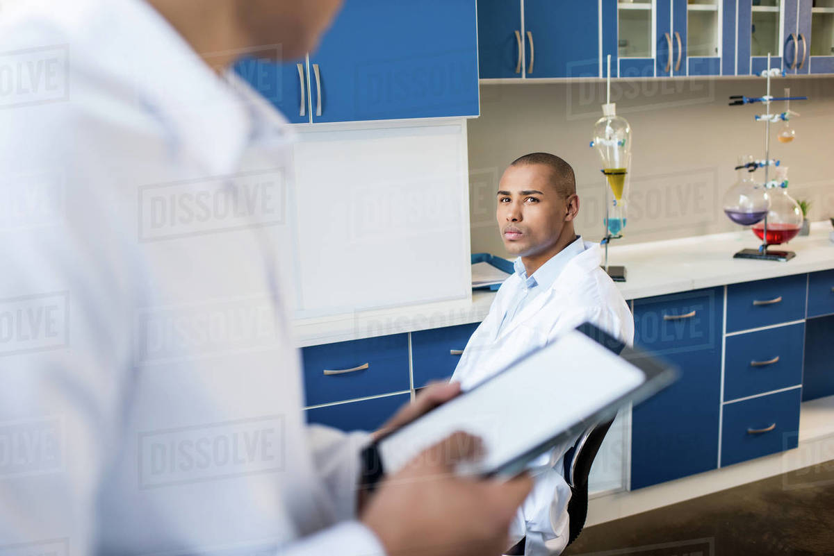 Young technician sitting in laboratory and talking to colleague holding ...