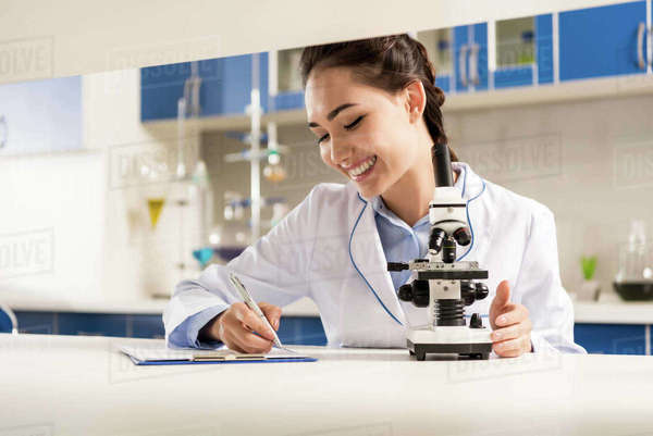 Young smiling lab technician taking notes after doing microscope sample ...