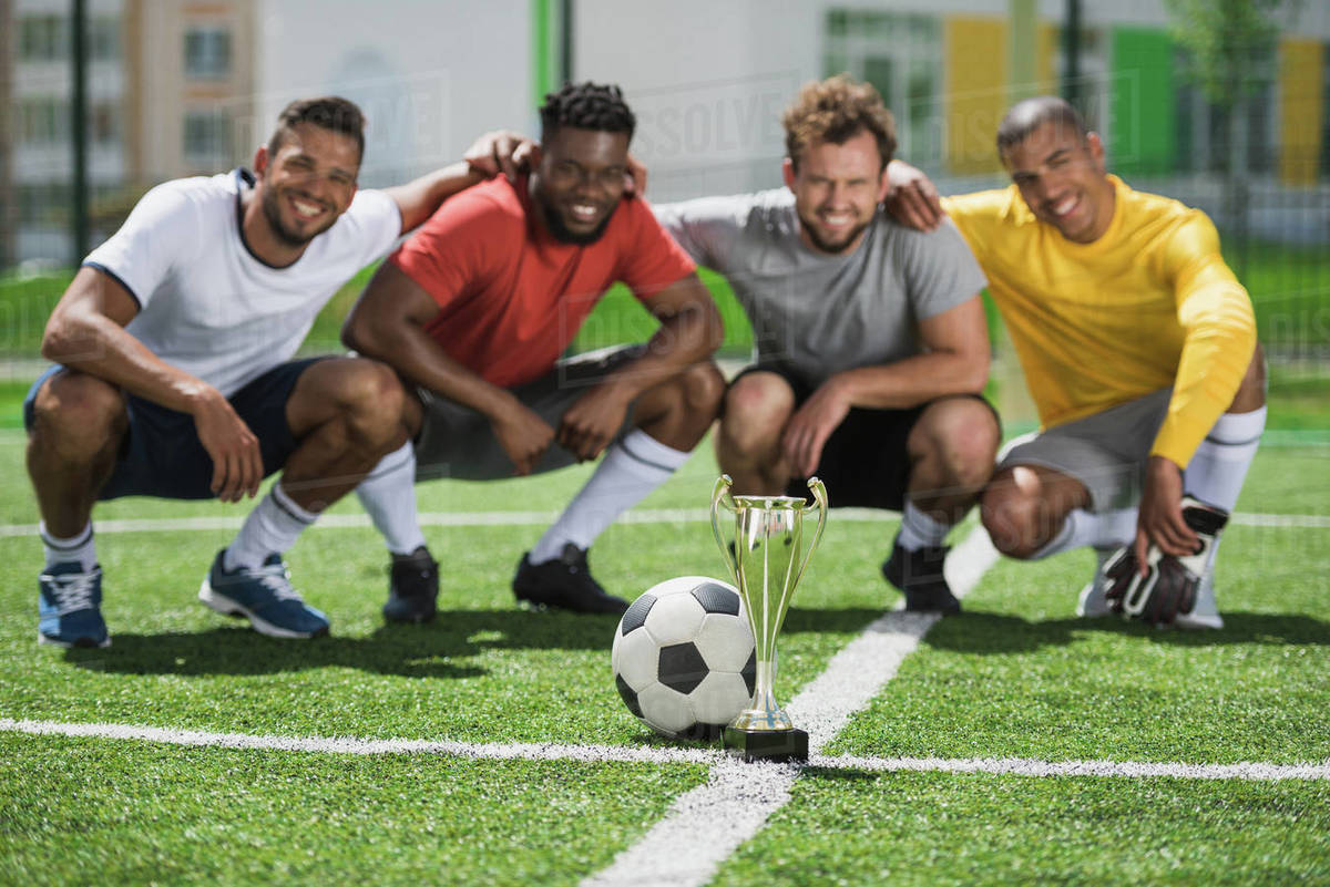 Smiling multiethnic soccer team with goblet on soccer pitch after game ...