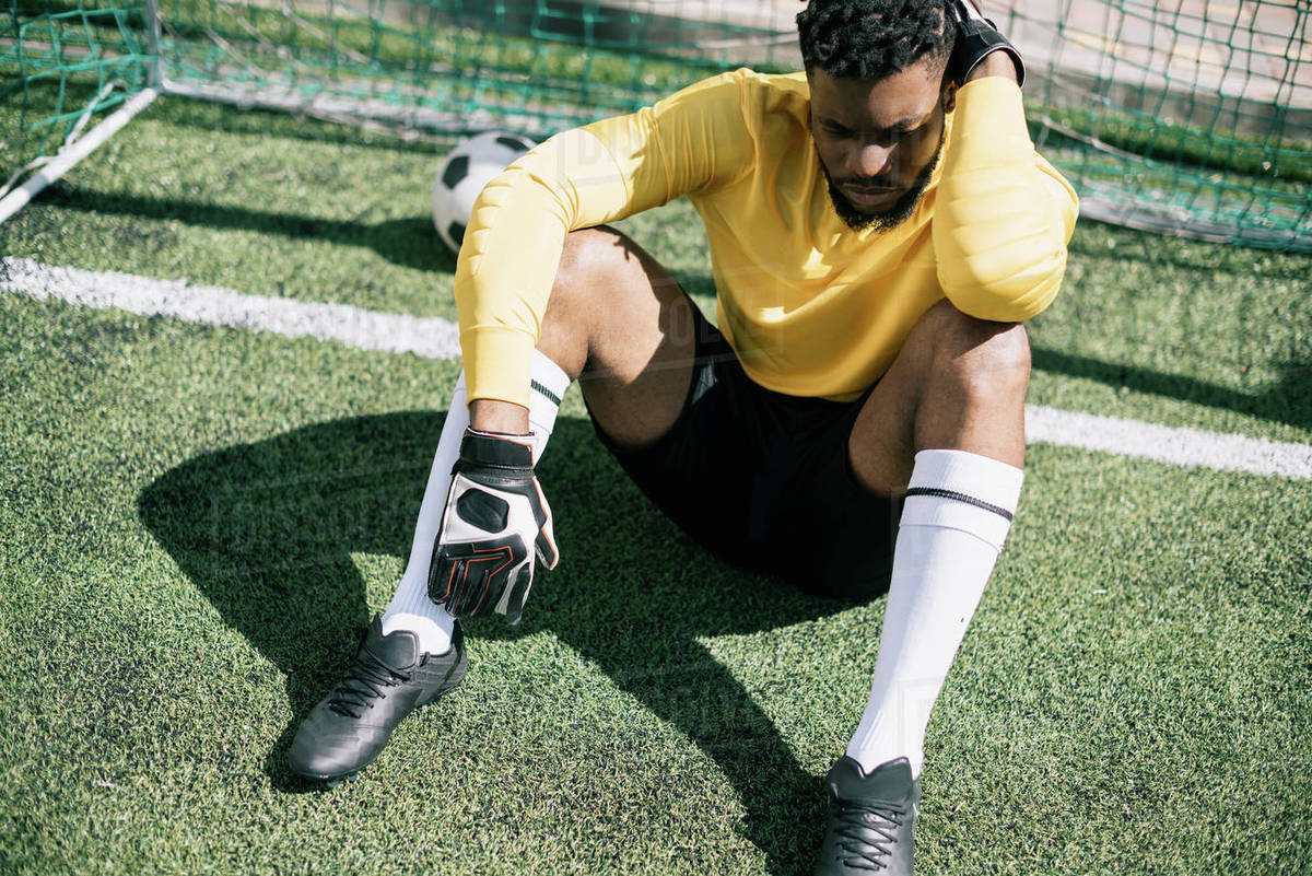 Upset african american goalkeeper sitting at goalpost on pitch ...