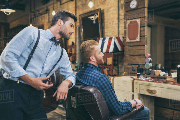 Stylish young barber with scissors standing behind customer that ...