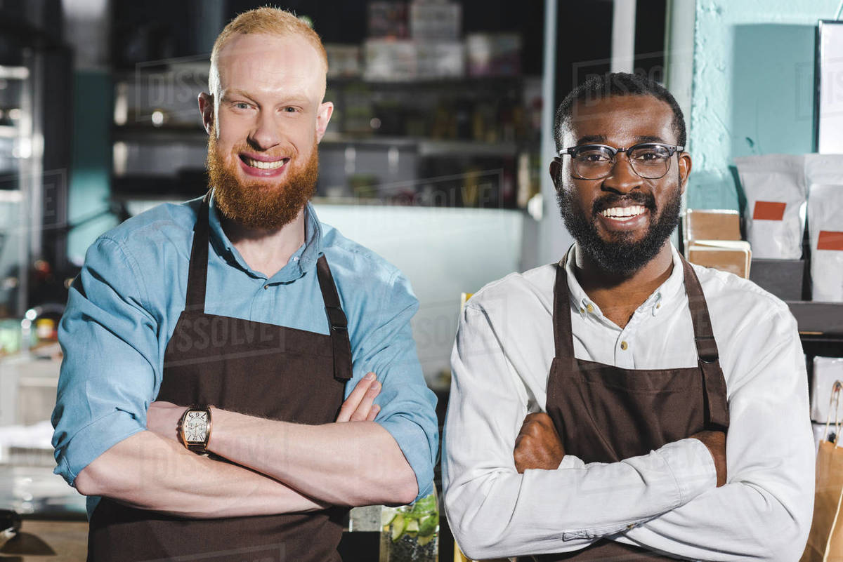 Portrait of two young multiethnic owners of coffee shop in aprons ...