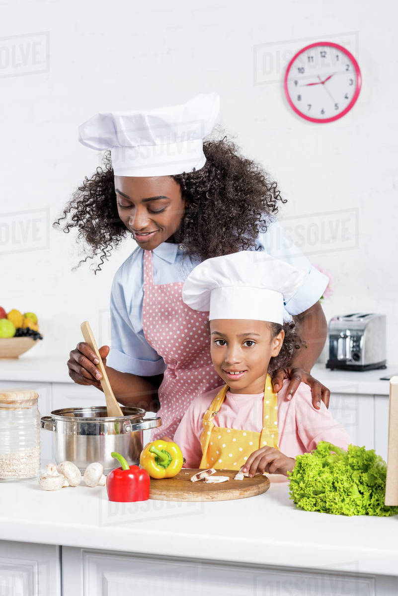 African american mother and daughter in chef hats cooking together on ...