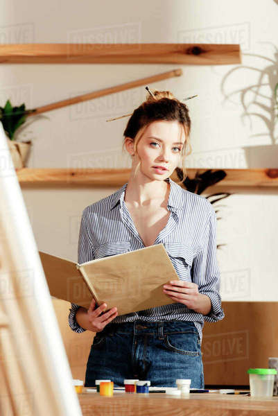 Attractive female artist reading book and standing at table with ...