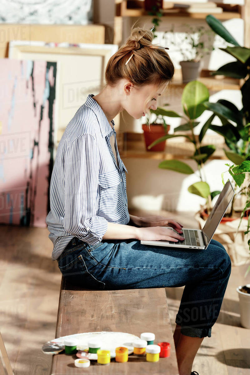 Side view of young female artist sitting on bench with paints and using ...