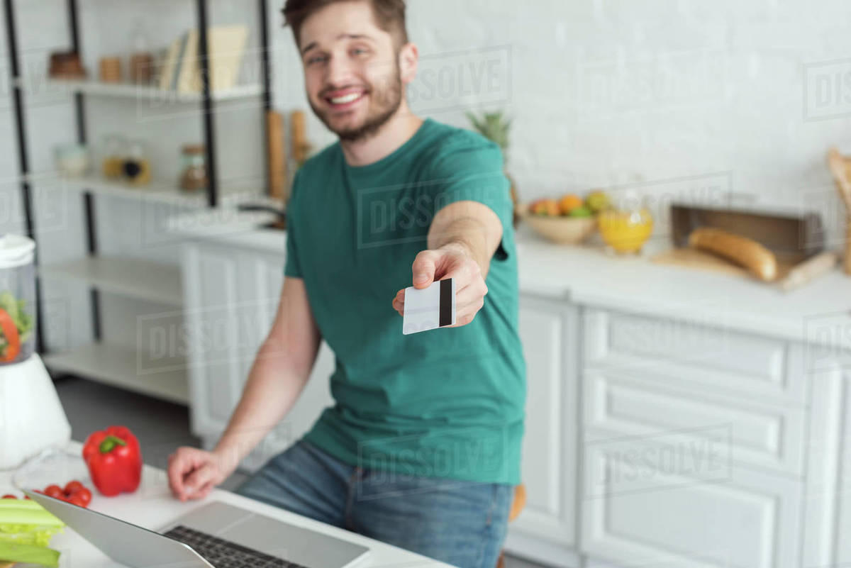 Smiling man showing credit card at table with laptop in kitchen at home ...